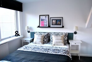 Quarto com cama, duas mesas de cabeceira, janela com persianas e duas imagens emolduradas na parede.