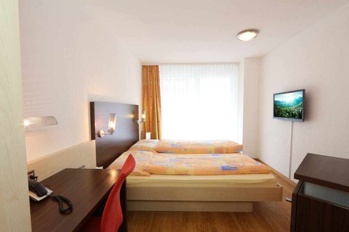Quarto de hotel com cama, escrivaninha com telefone, cadeira, televisão e janela com cortinas.