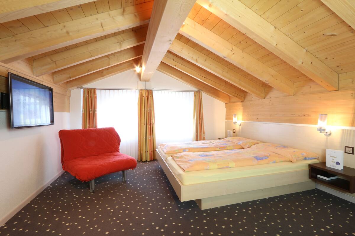 Quarto com teto de madeira, uma cama, uma cadeira vermelha e uma janela com cortinas.