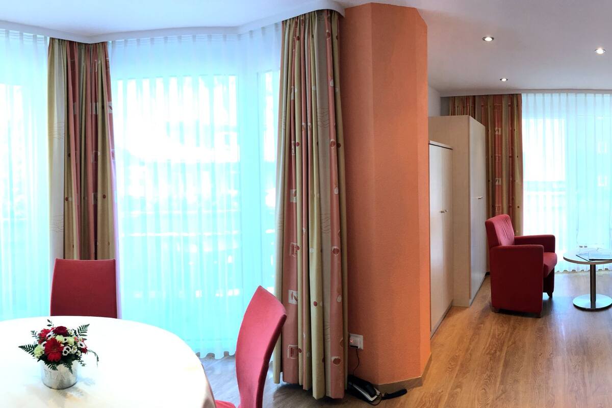 Quarto de hotel moderno com cozinha, mesa de jantar com cadeiras e um pequeno espaço de estar com uma cadeira vermelha.