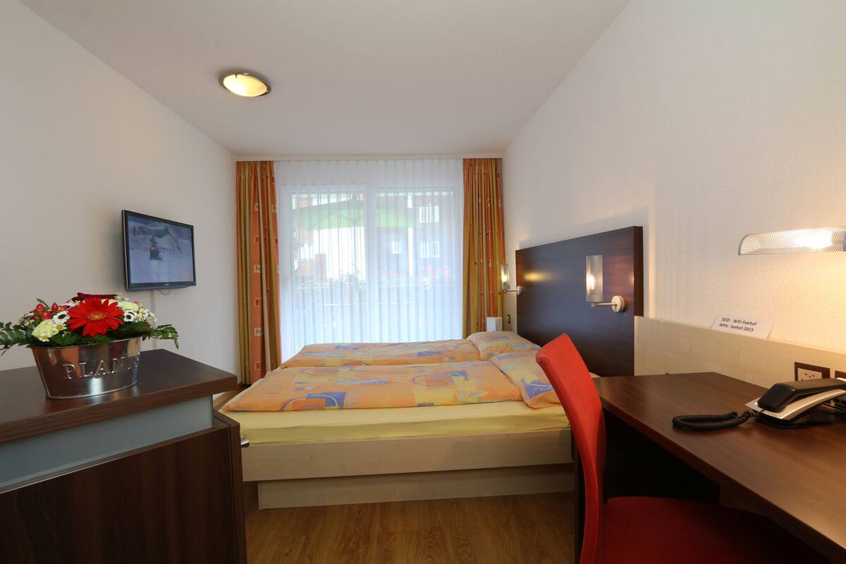 Quarto de hotel com cama, escrivaninha, cadeira, televisão, telefone e sinal indicando acesso à rede Wi-Fi.