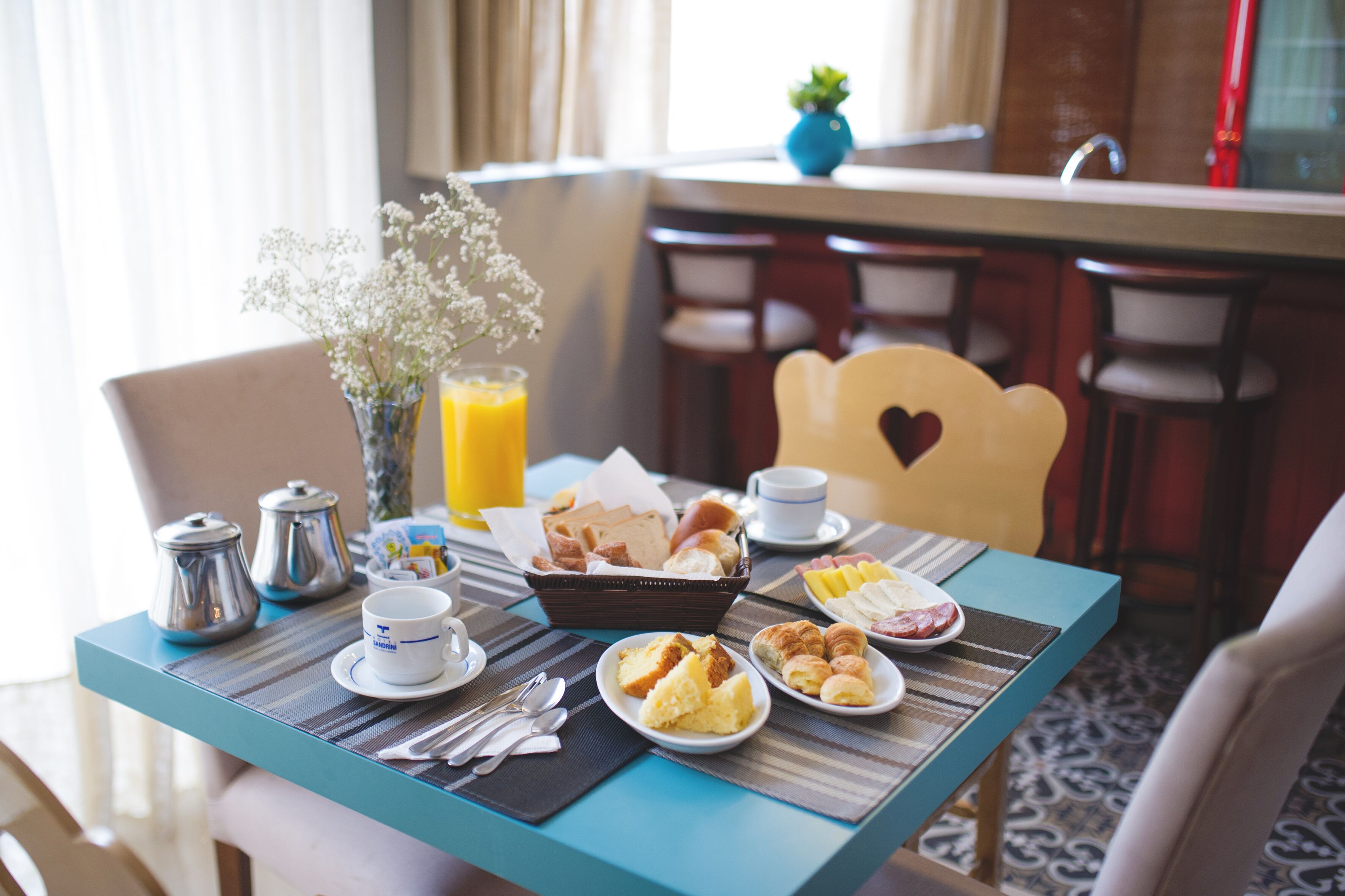 Mesa de café da manhã com diversos alimentos, uma cafeteira, xícaras e um vaso com flores.