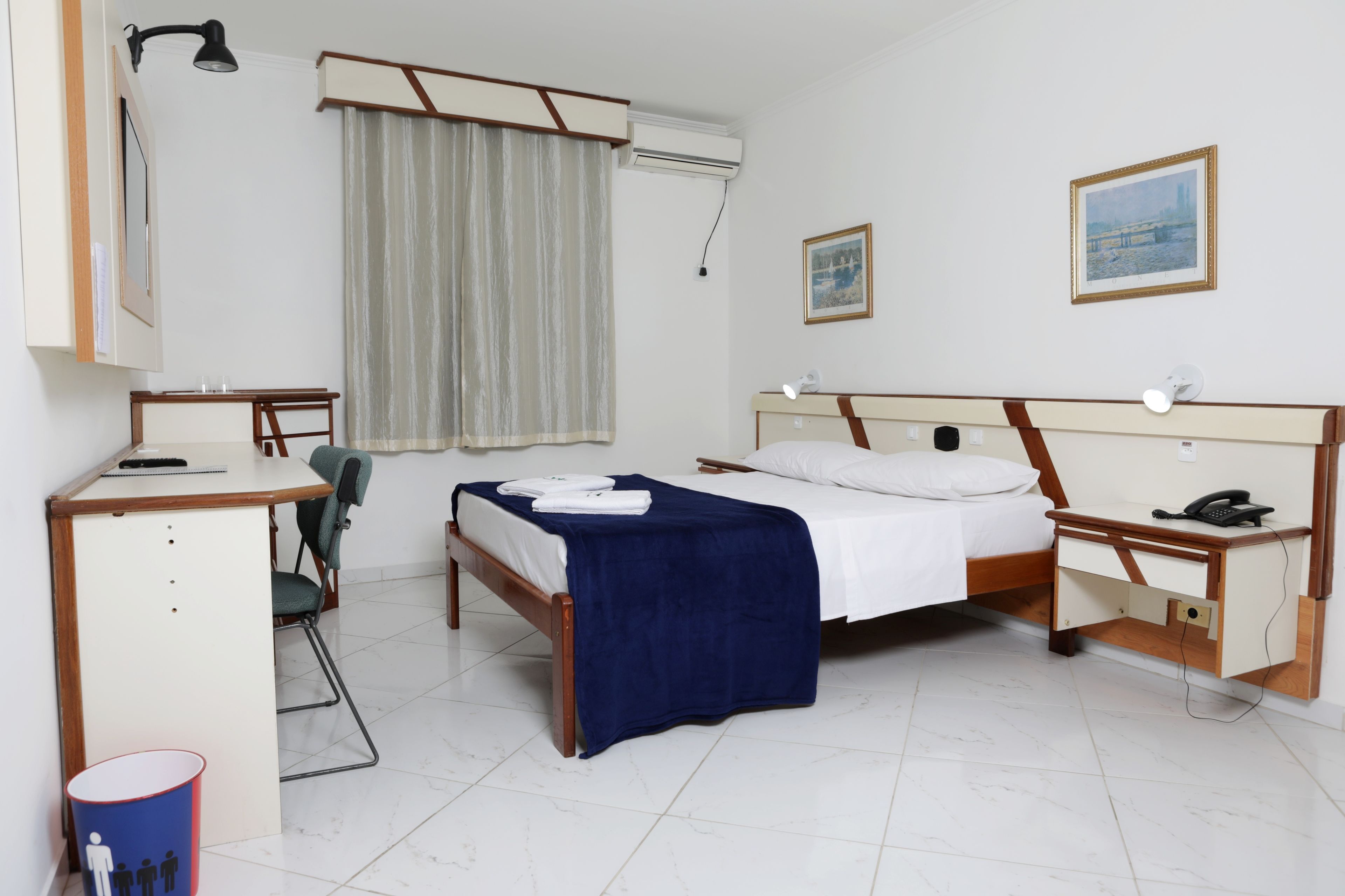 Quarto de hotel com cama, escrivaninha, cadeira e uma mesinha de cabeceira com abajur.