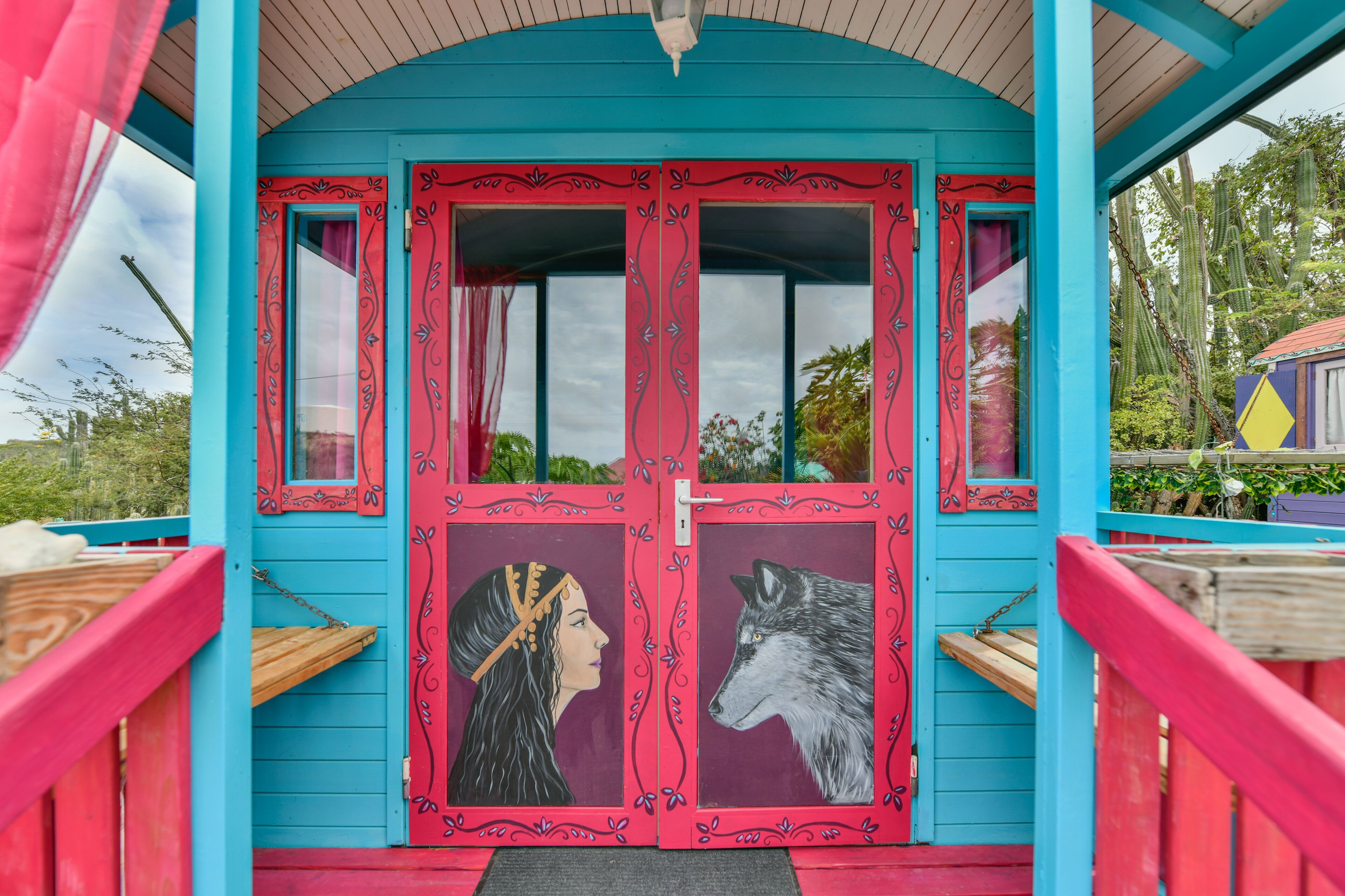 Um edifício colorido com telhado turquesa e portas vermelhas, apresentando um mural de uma mulher e um lobo.