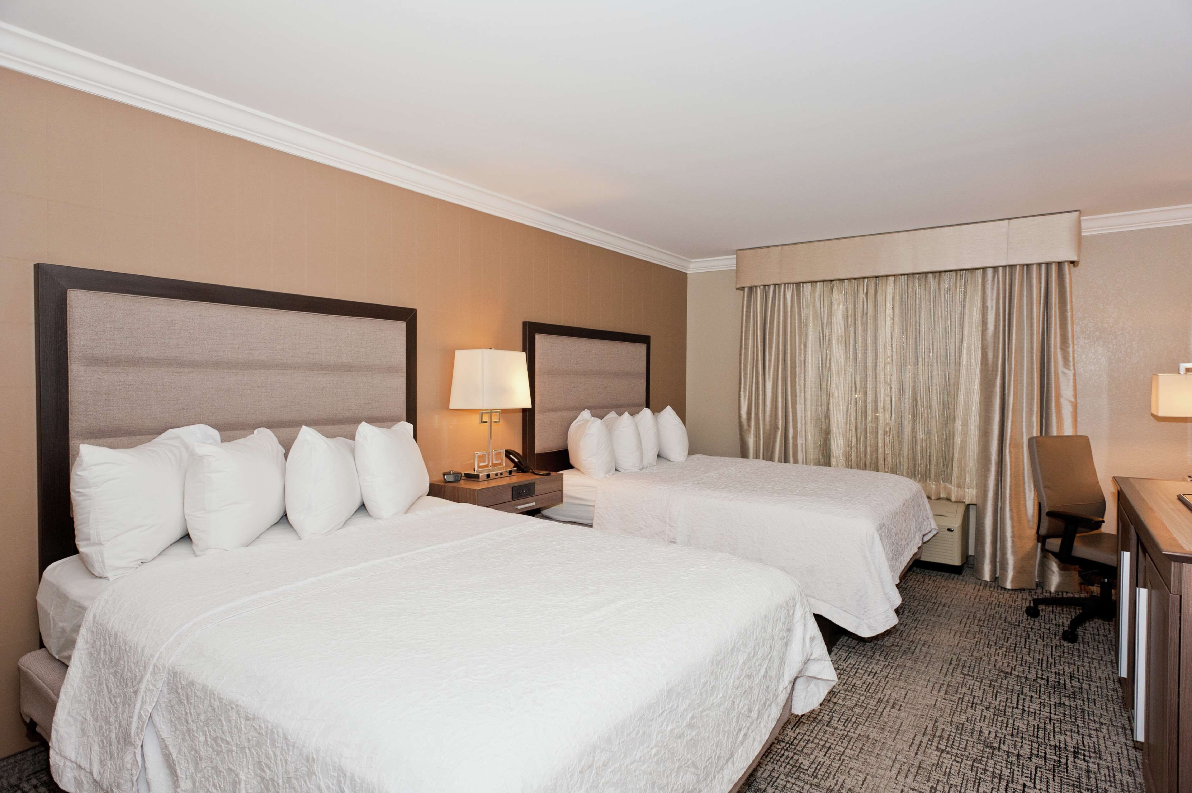 Quarto de hotel com duas camas, uma escrivaninha, uma cadeira e uma janela grande com cortinas.