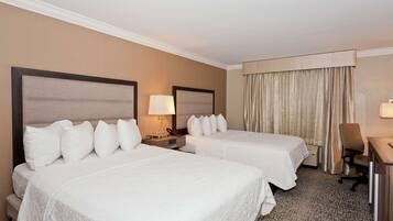 Quarto de hotel com duas camas, uma escrivaninha, uma cadeira e uma janela grande com cortinas.
