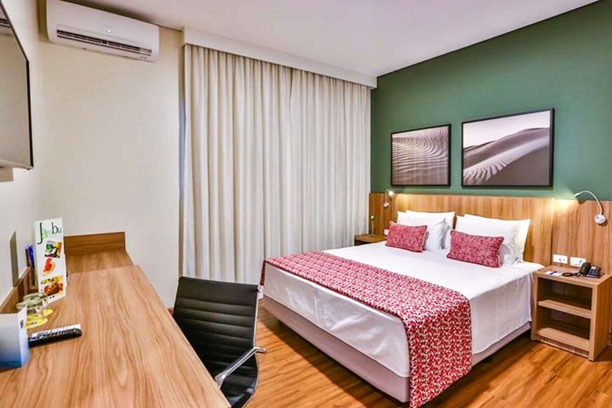Quarto de hotel com cama, escrivaninha, cadeira, mesa de cabeceira e duas obras de arte na parede.