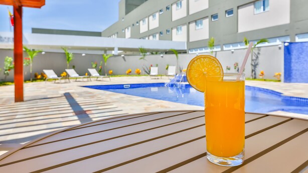 Um copo de suco de laranja sobre uma mesa, perto de uma piscina com espreguiçadeiras e um edifício ao fundo.