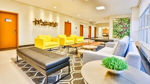 Um hall de hotel moderno com área de estar, com cadeiras amarelas, um sofá cinza e um banco preto. Há uma peça decorativa na parede acima da área de estar e uma janela com vista para vegetação.