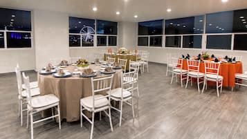 Uma sala de banquetes com mesas redondas preparadas para um evento formal, com cadeiras brancas e mesas cobertas por toalhas bege.