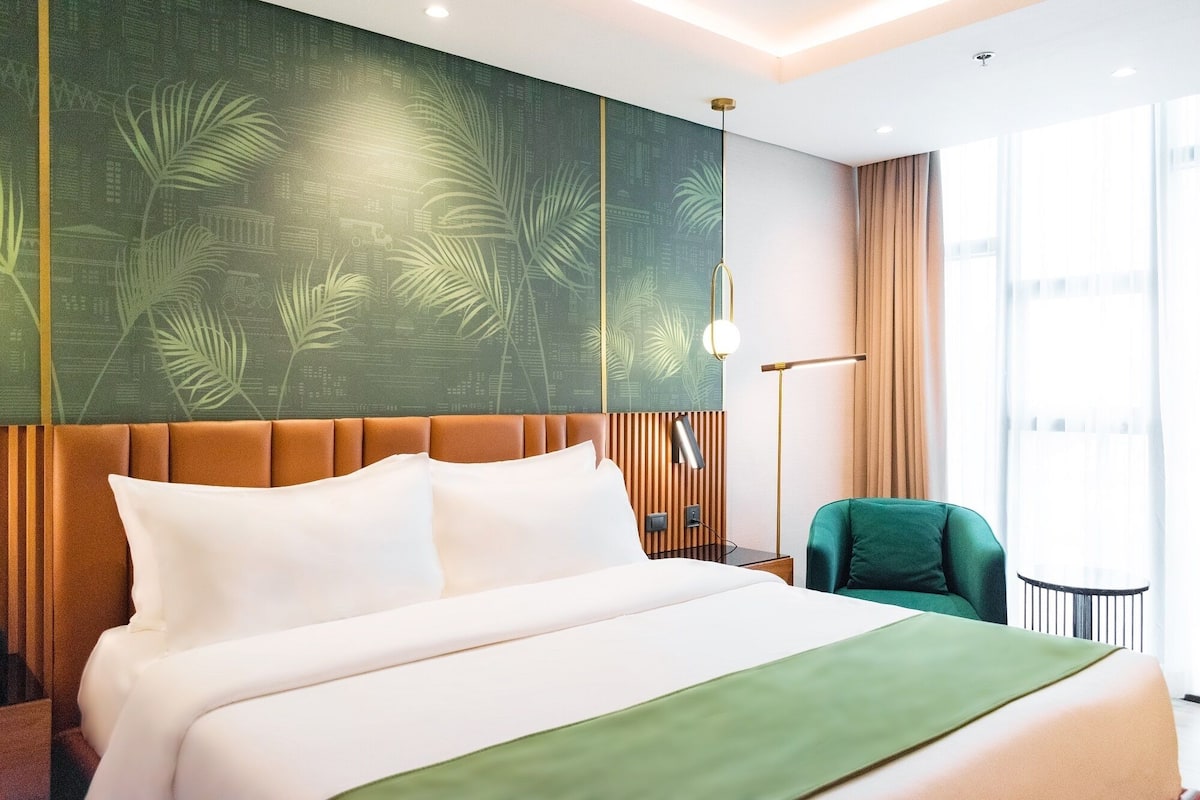 Quarto de hotel com uma cama grande, uma poltrona verde e papel de parede com vista para a cidade.