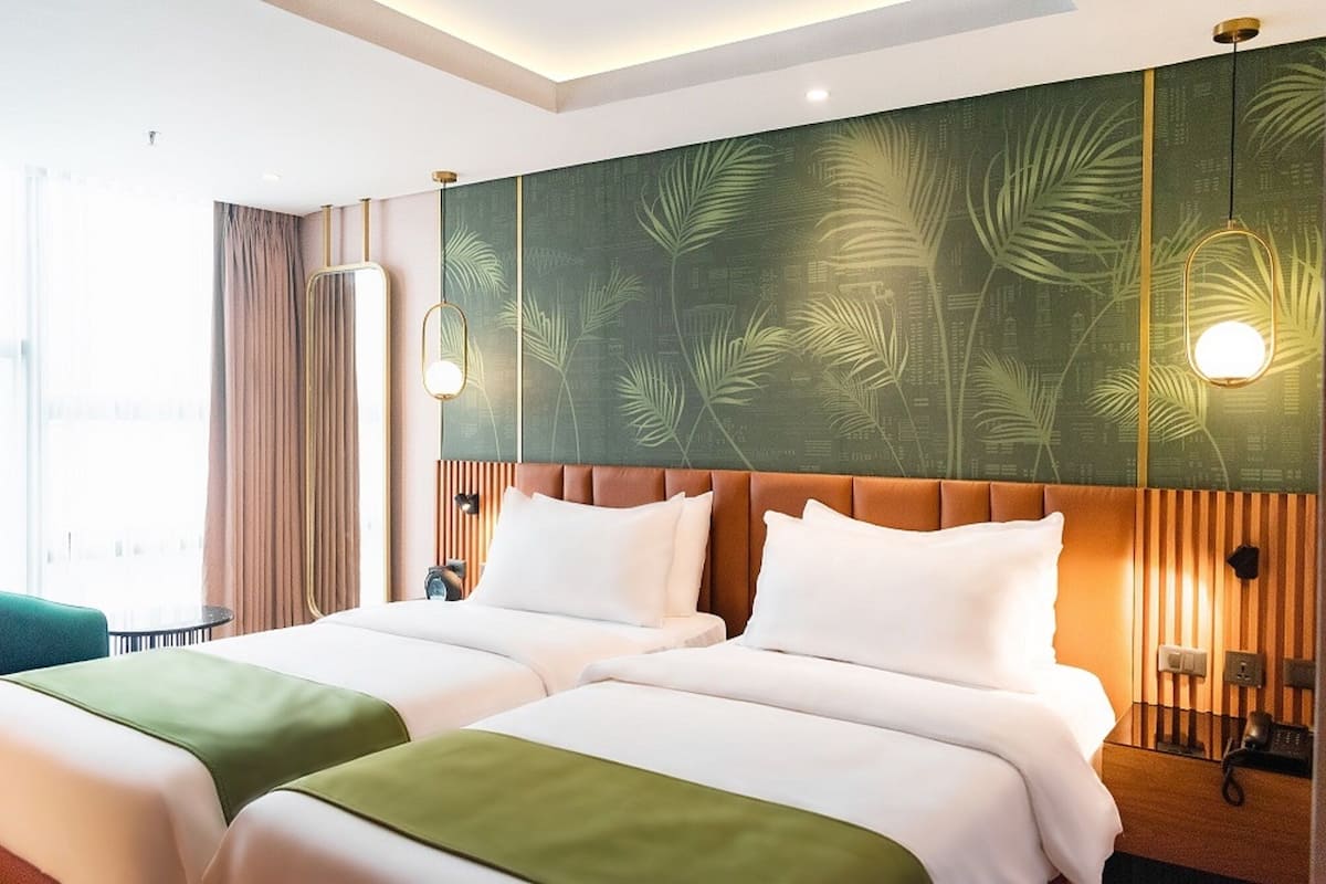 Quarto de hotel com duas camas, cabeceira estampada com folhagens, mesa de cabeceira com luminária e janela com cortinas.
