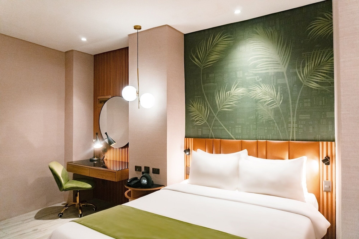 Quarto de hotel com cama, secretária e cadeira, espelho e parede com padrão de folha.