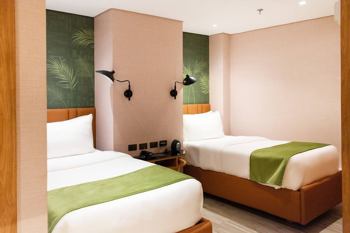 Quarto de hotel com duas camas, uma mesa de cabeceira, um relógio e uma luminária na parede.