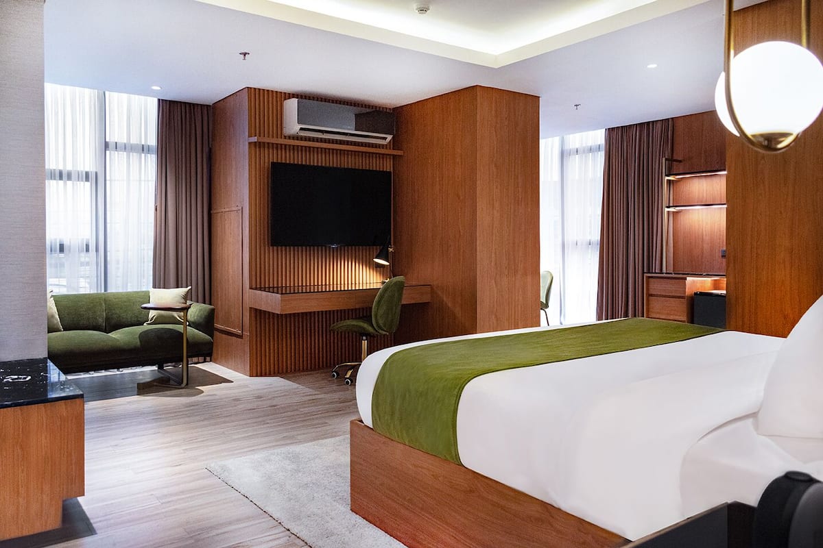 Quarto de hotel moderno com uma cama grande, uma escrivaninha com cadeira, uma televisão e uma pequena área de estar com sofá e poltrona.