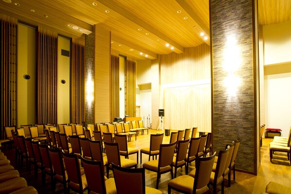 Sala de conferências com fileiras de cadeiras, palco e parede de pedra decorativa.