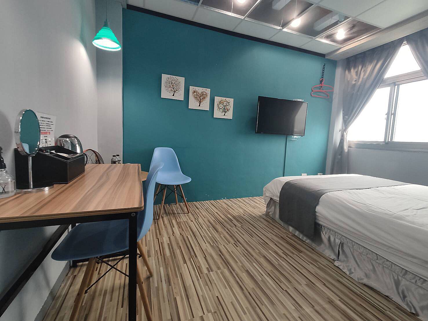 Quarto de hotel com piso de madeira, uma cama, uma escrivaninha com cadeira, uma televisão fixada na parede e uma janela com cortinas.