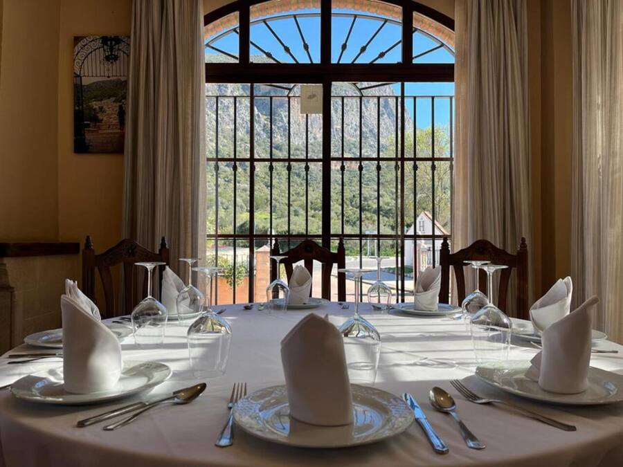 Mesa de jantar posta para seis pessoas, com vista para uma paisagem montanhosa através de uma grande janela com grades de metal.