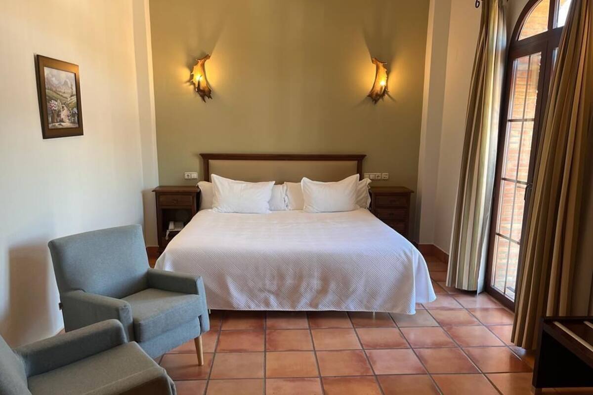 Quarto de hotel com uma cama grande, duas mesas de cabeceira, uma cadeira e uma janela com cortinas.