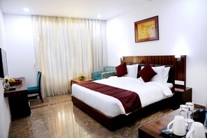 Quarto de hotel com uma cama grande, cabeceira de madeira, escrivaninha com cadeira, mesinha com um vaso e um quadro na parede.