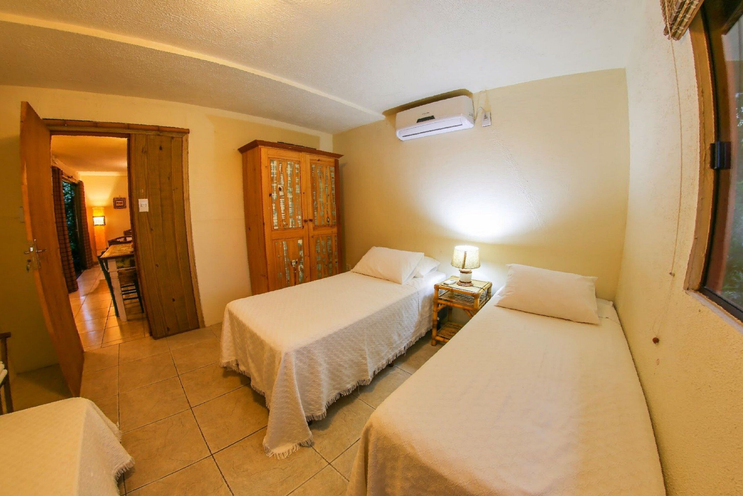 Quarto com duas camas, guarda-roupa de madeira, mesa de cabeceira com abajur e ar condicionado.