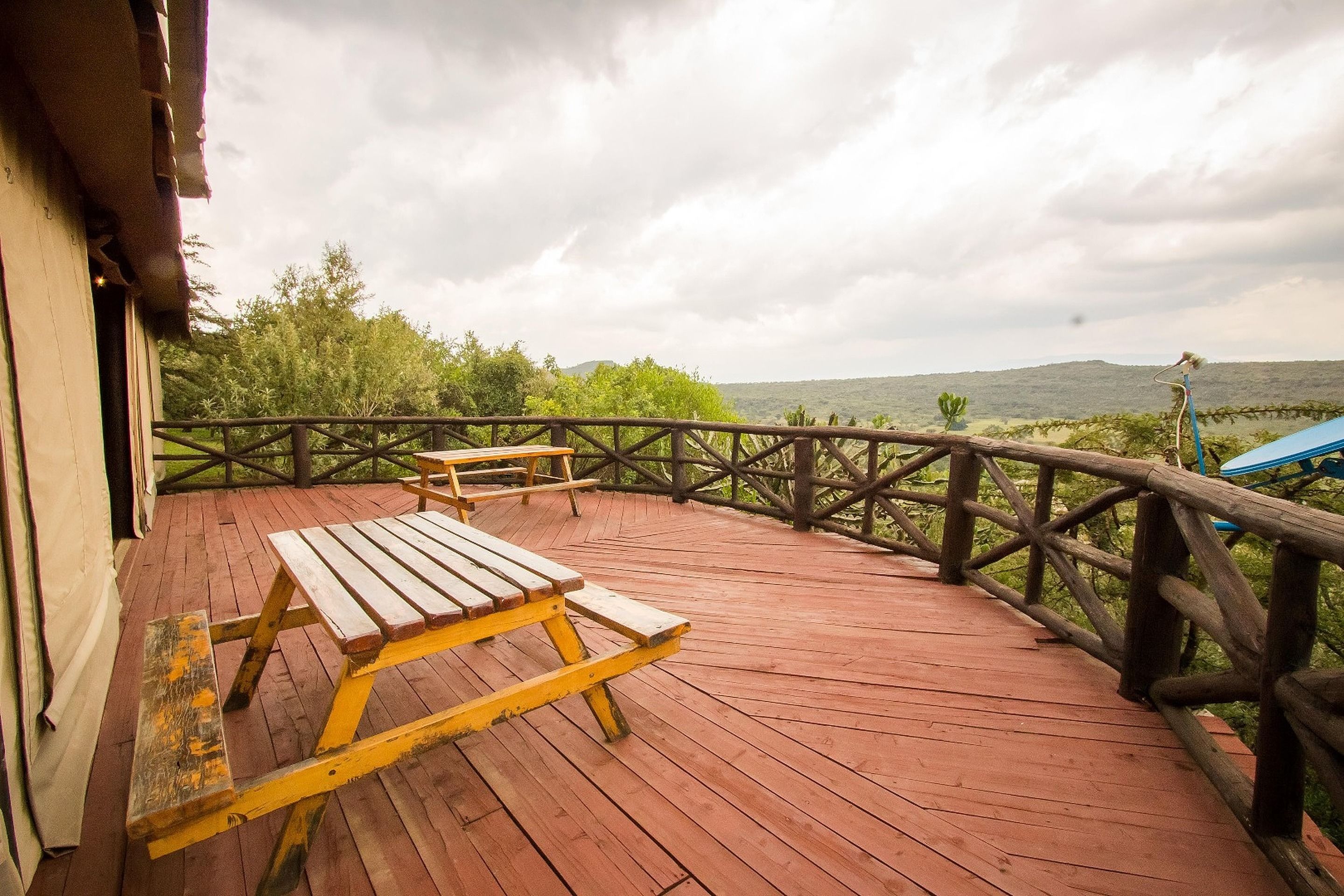 Um deck de madeira com mesas de piquenique e guarda-corpo, com vista para uma paisagem natural.
