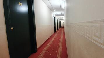 Um corredor com carpete vermelho, paredes brancas e uma porta preta com placa de número.