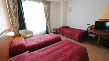 Quarto de hotel com duas camas de solteiro, uma mesa redonda pequena, uma cadeira, televisão e janela com cortinas.
