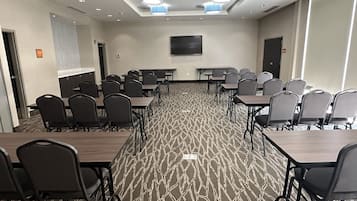Uma sala de conferência com carpete estampado, várias mesas e cadeiras, uma TV de tela plana e um candeeiro no teto.