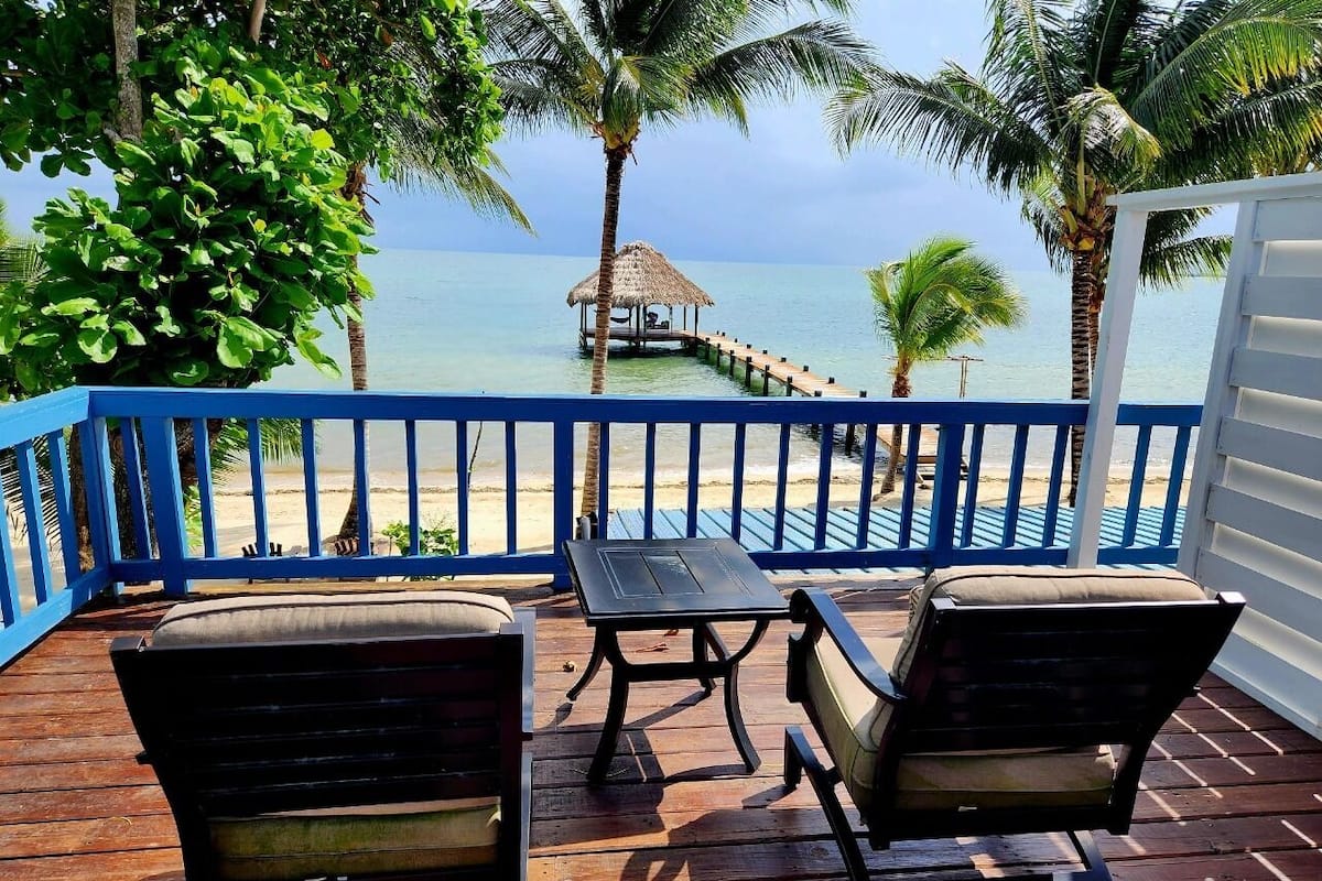 Um deck de madeira com mesa e cadeiras, com vista para uma praia com uma estrutura de palha e palmeiras.