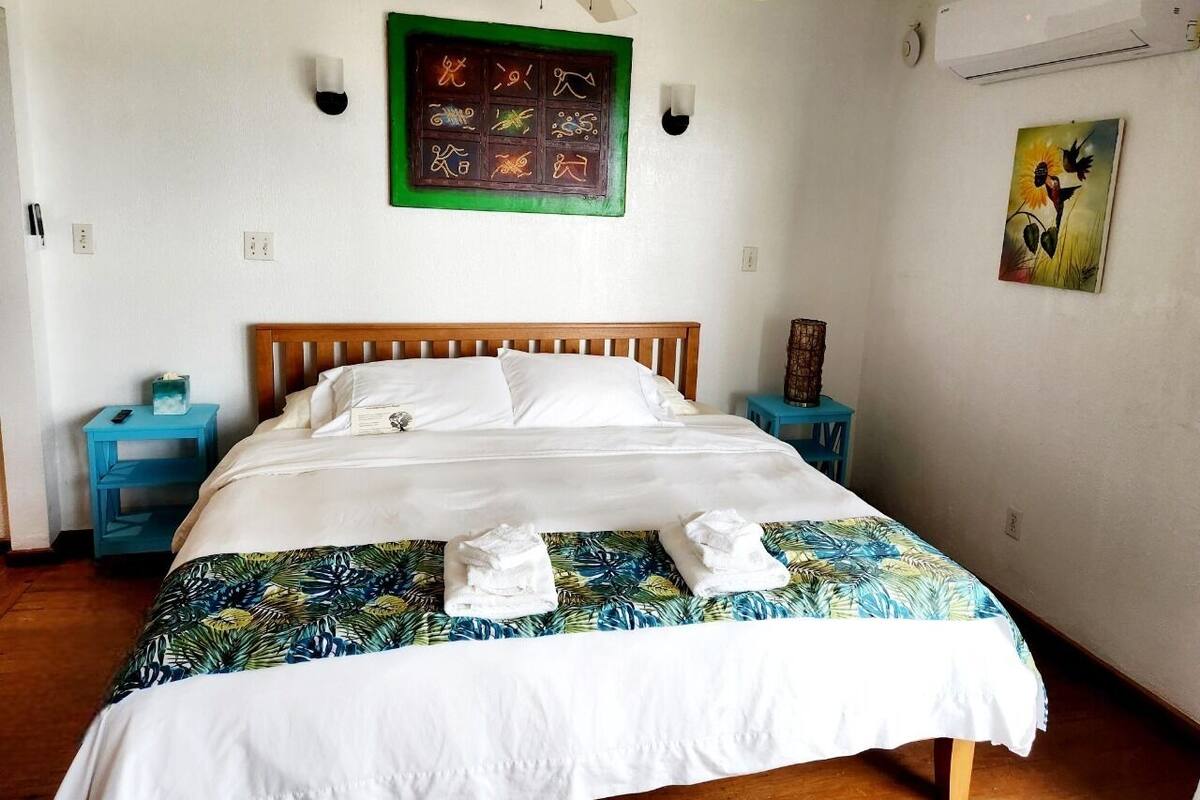 Uma cama bem arrumada com colcha florida, um ventilador de teto, uma obra de arte na parede e uma mesinha com abajur.