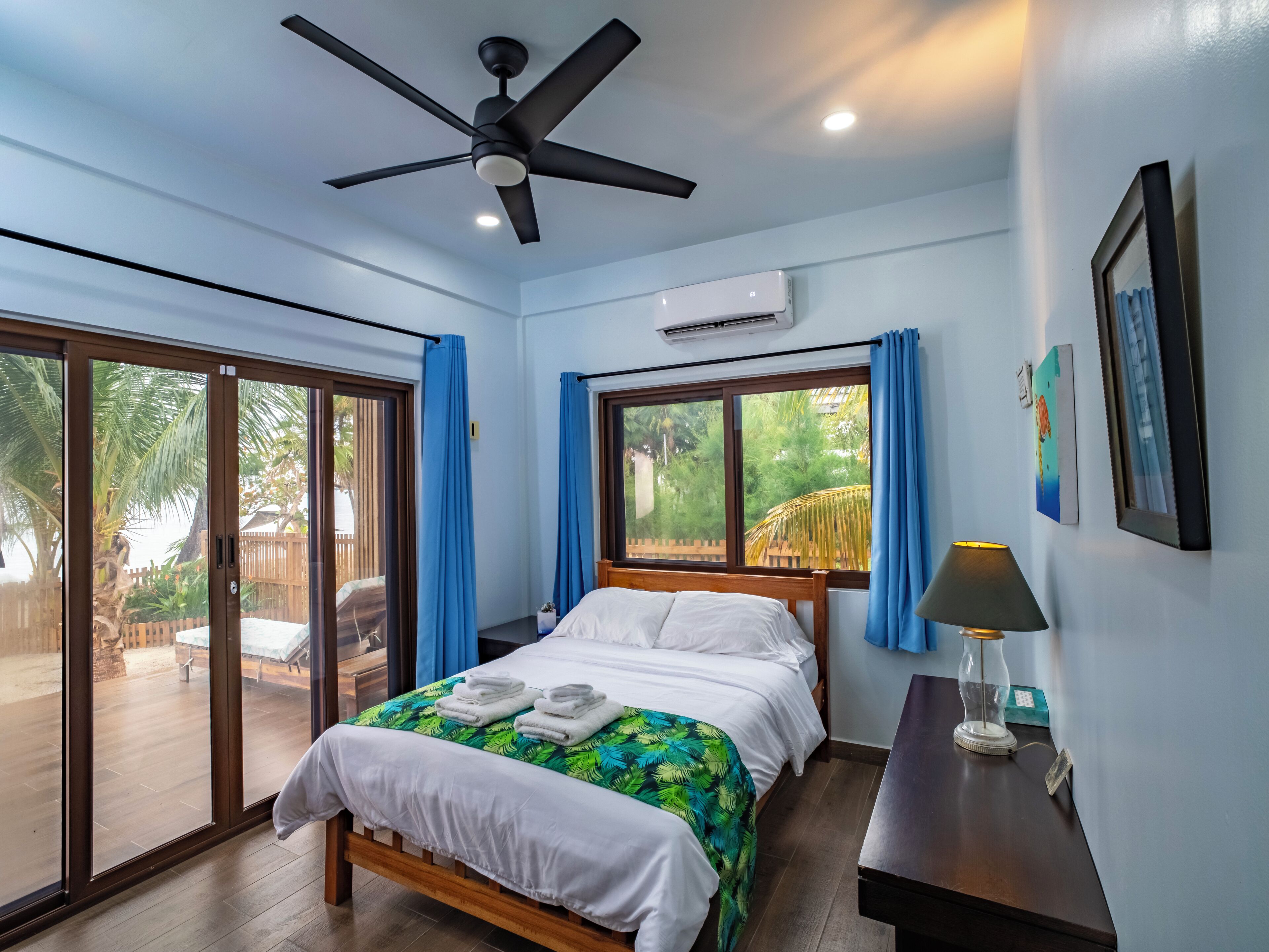 Quarto com cama, ventilador de teto, ar condicionado, mesa de cabeceira, abajur e vista para o exterior.