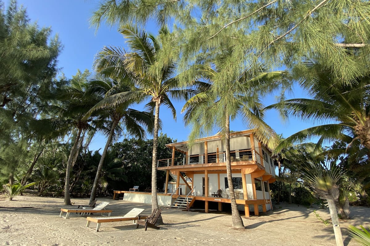 Uma casa de praia de dois andares com varanda, rodeada por palmeiras.