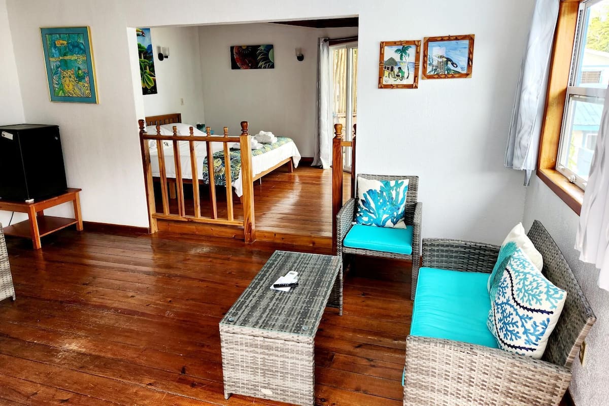 Uma sala de estar aconchegante com piso de madeira, uma cama com cabeceira de madeira, um sofá de vime com almofadas azuis e uma televisão sobre uma mesinha.