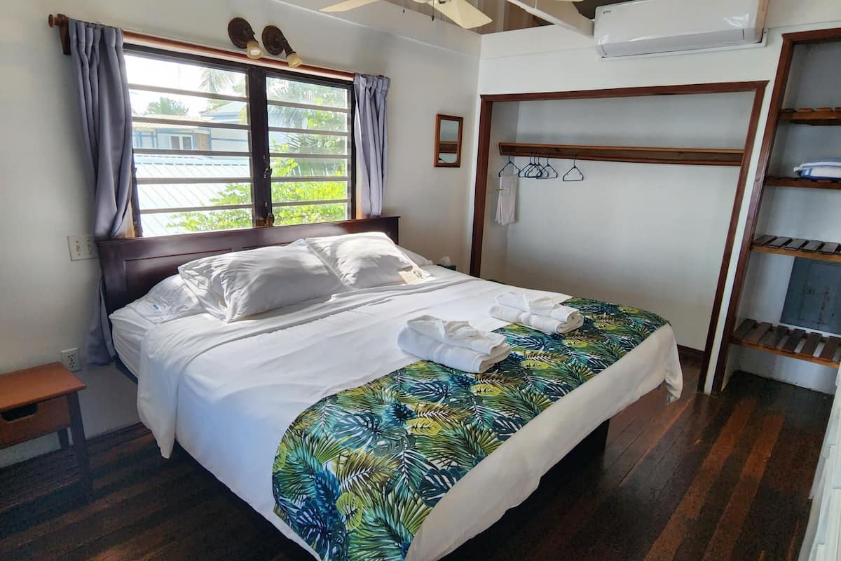 Cama bem arrumada com lençóis brancos e um edredom com padrão de folhas tropicais, mesa de cabeceira de madeira, ventilador de teto e janela com cortinas.