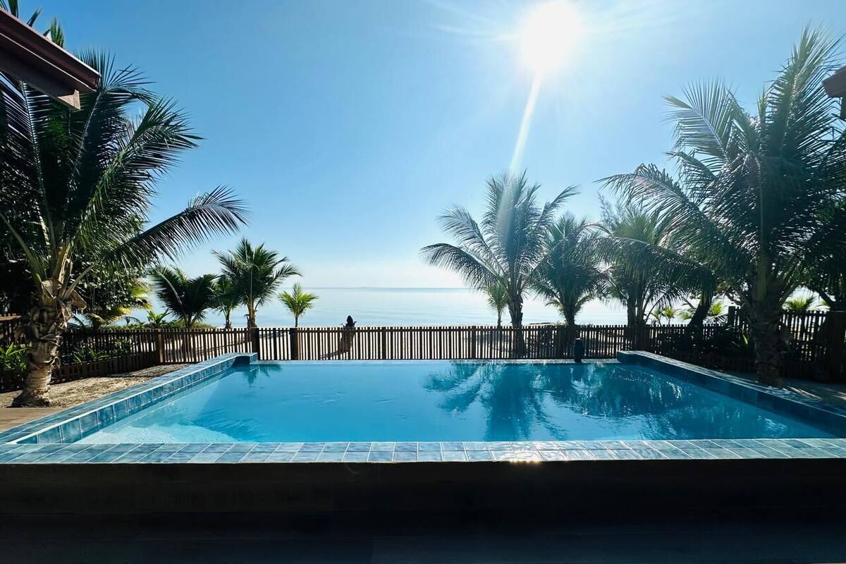 Uma piscina retangular com água azul cristalina, rodeada por palmeiras e um cercado de madeira, com vista para o oceano ao fundo.
