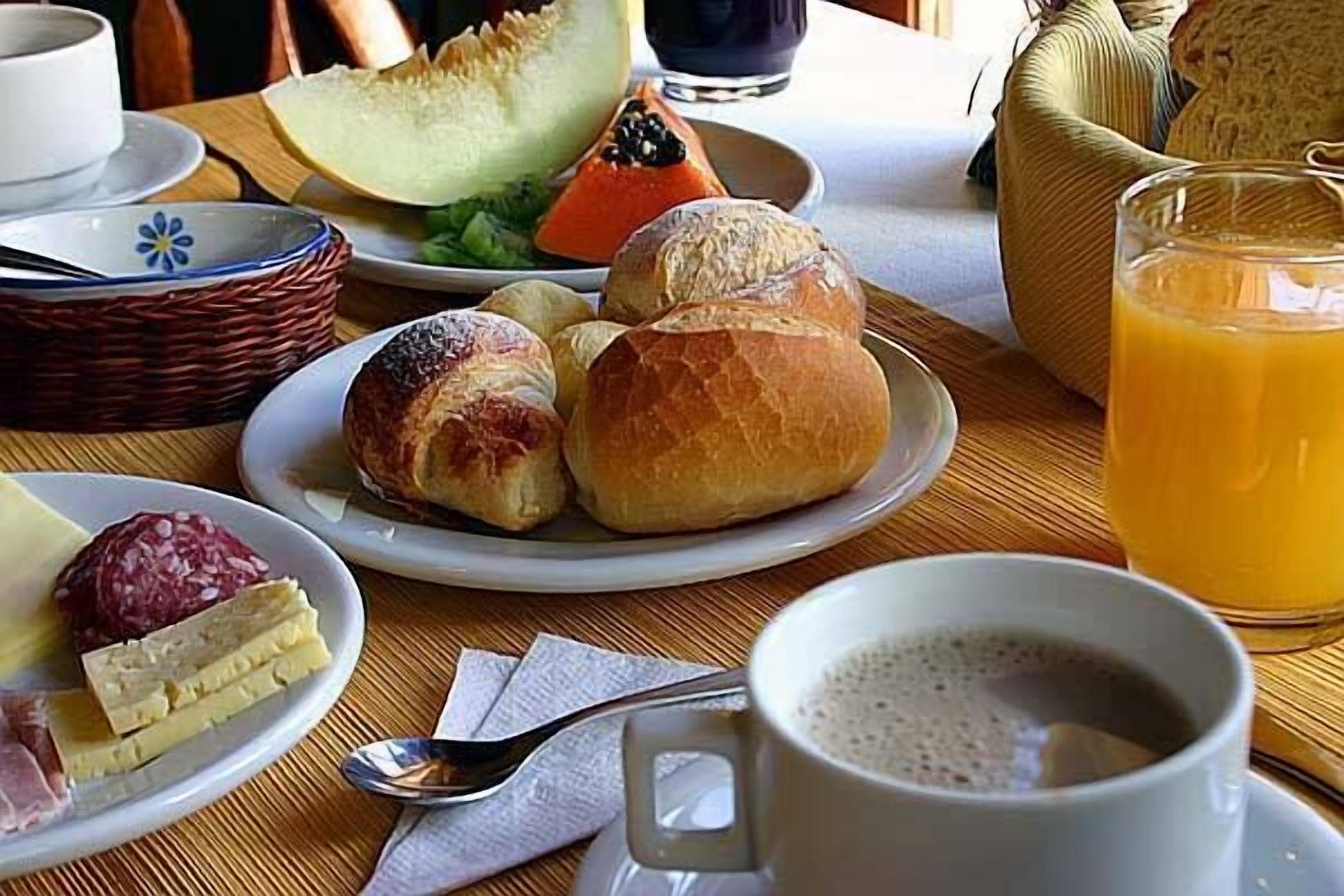 Mesa de café da manhã com croissants, queijo, salada de frutas e um copo de suco de laranja.