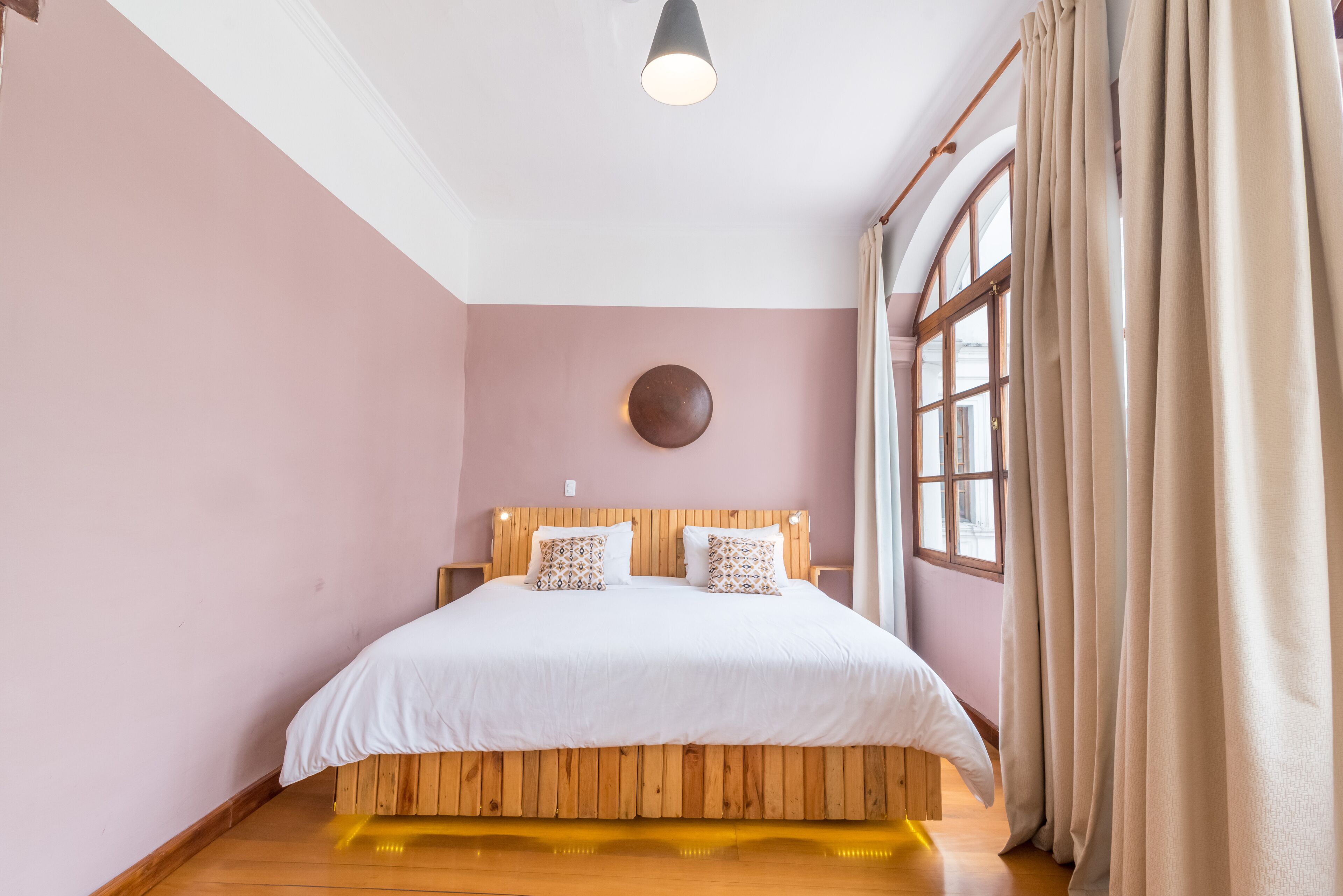 Quarto com cabeceira de madeira, cama grande com roupa de cama branca, relógio de parede redondo e janela com cortinas.