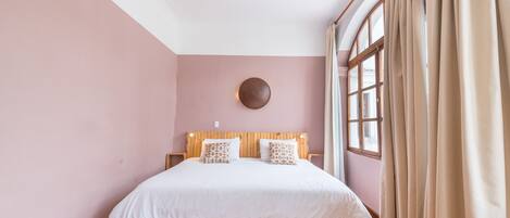 Quarto com cabeceira de madeira, cama grande com roupa de cama branca, relógio de parede redondo e janela com cortinas.