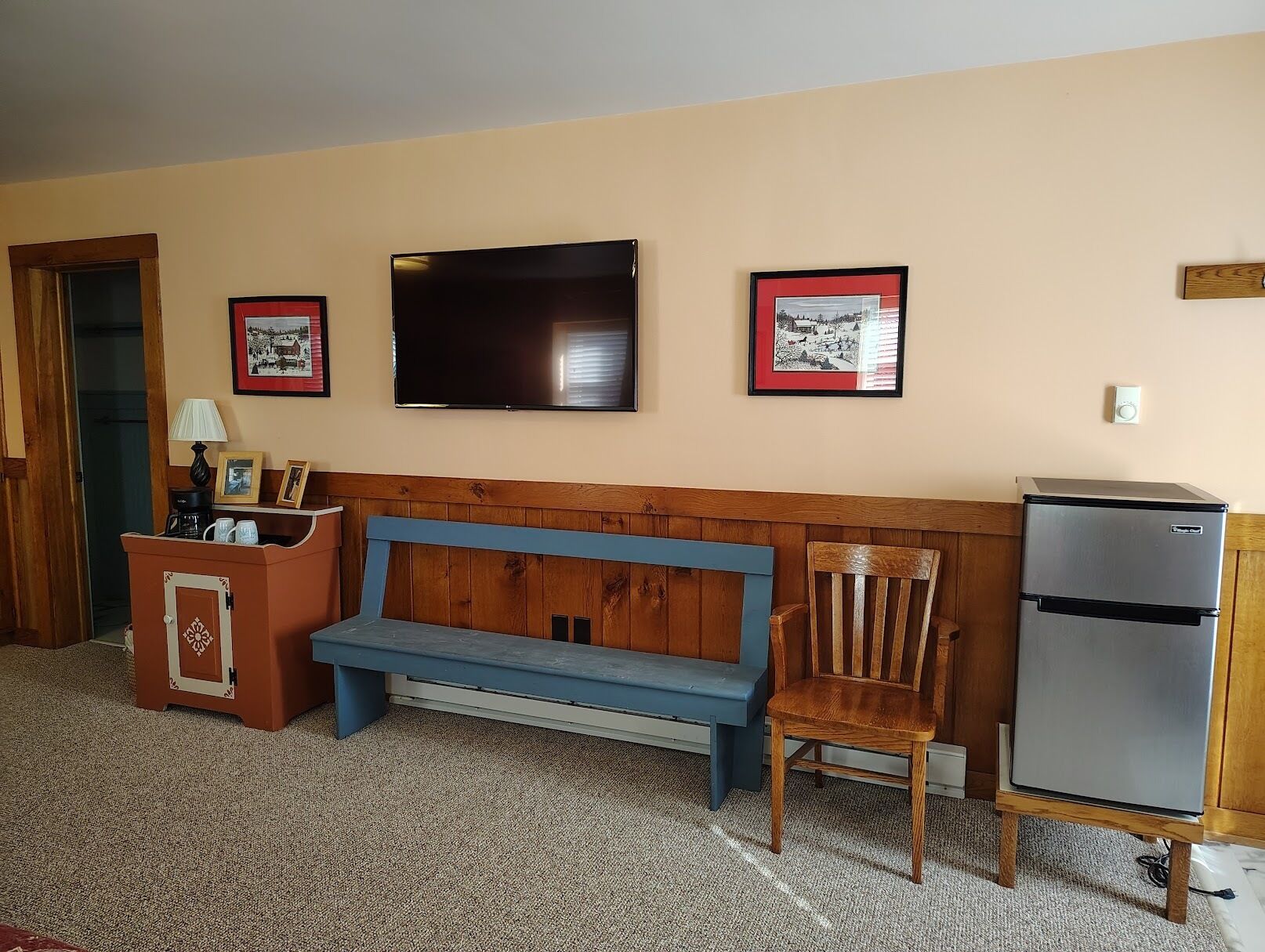 Quarto com um banco de madeira, uma mesa pequena com uma luminária, um refrigerador e duas imagens emolduradas na parede.