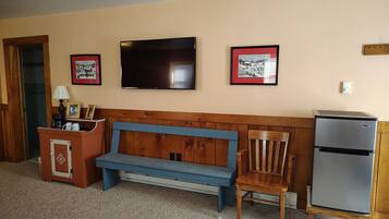 Quarto com um banco de madeira, uma mesa pequena com uma luminária, um refrigerador e duas imagens emolduradas na parede.