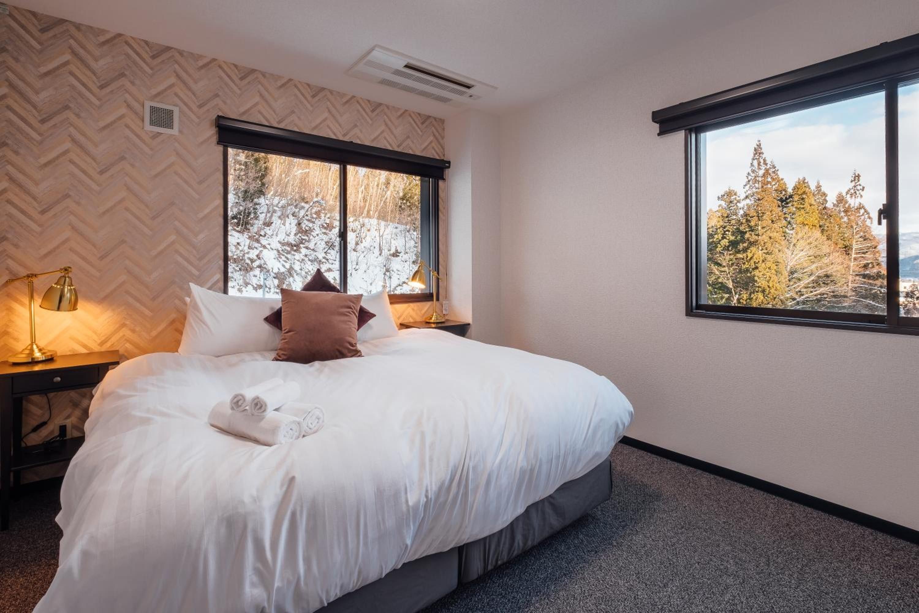 Quarto de hotel com uma cama grande, duas mesas de cabeceira, uma janela com vista para árvores e uma parede com padrão losango.