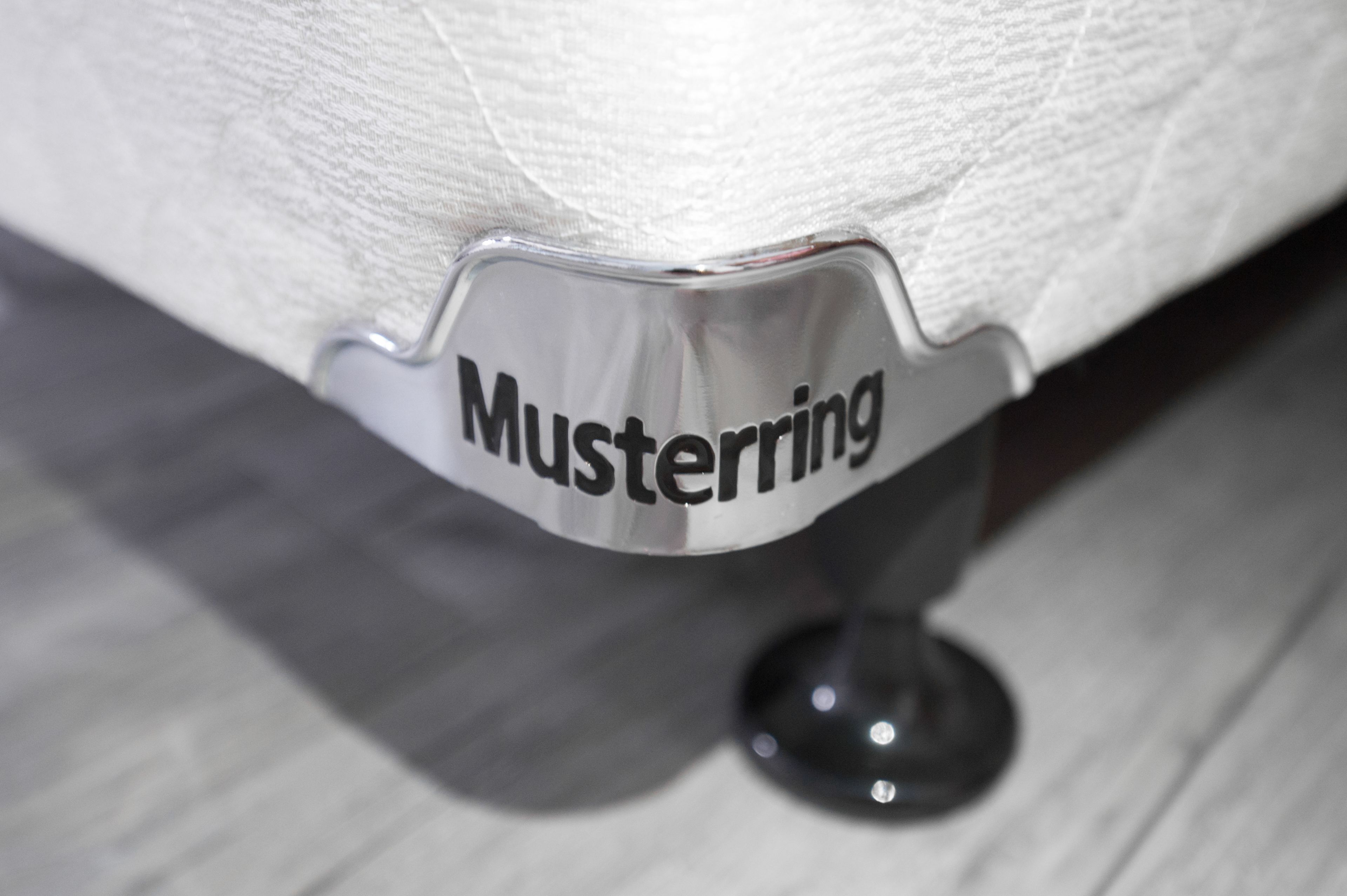 Um close-up de um objeto metálico com a palavra 'Mustering' gravada, preso a uma superfície texturizada.