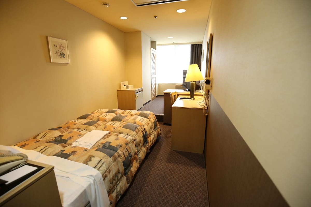 Quarto de hotel com cama, escrivaninha, cadeira, abajur e uma pequena janela.