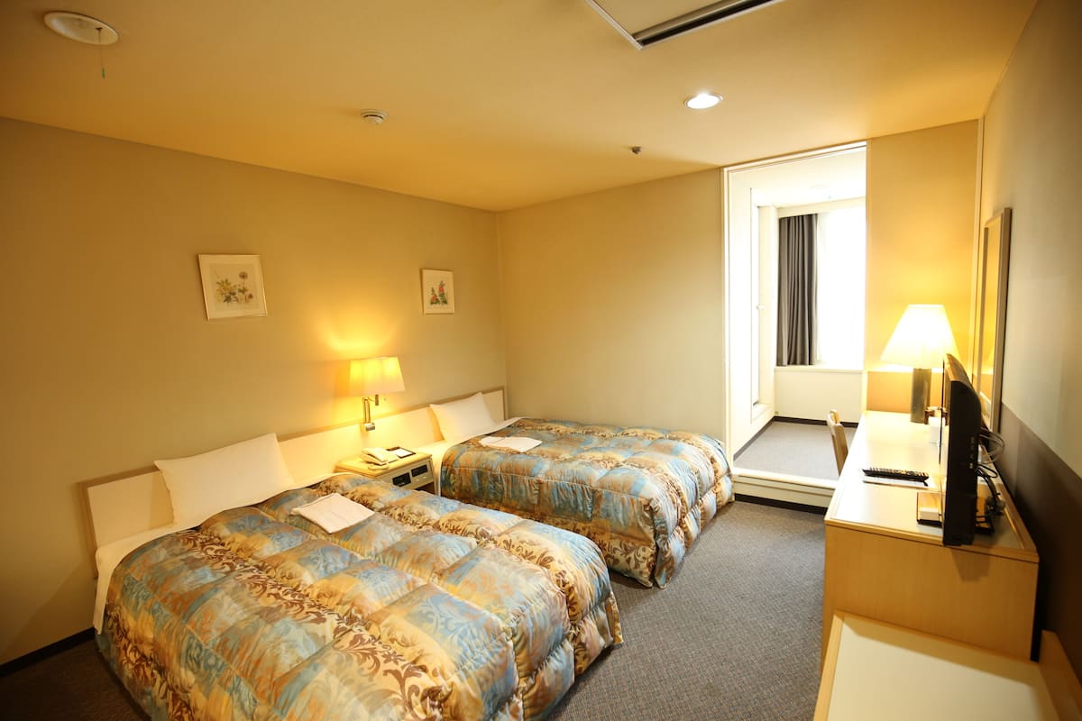 Quarto de hotel com duas camas, uma escrivaninha com computador e uma janela com cortinas.
