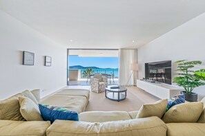 Uma sala de estar moderna com um sofá, uma mesa de centro redonda e uma TV. Há uma varanda com vista para o mar e as montanhas.