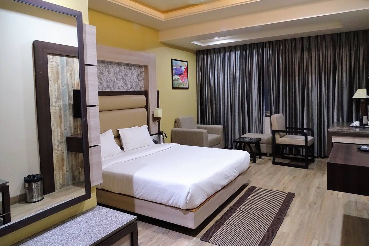 Quarto de hotel com uma cama grande, uma escrivaninha, duas cadeiras, uma mesinha, um abajur e um quadro na parede.