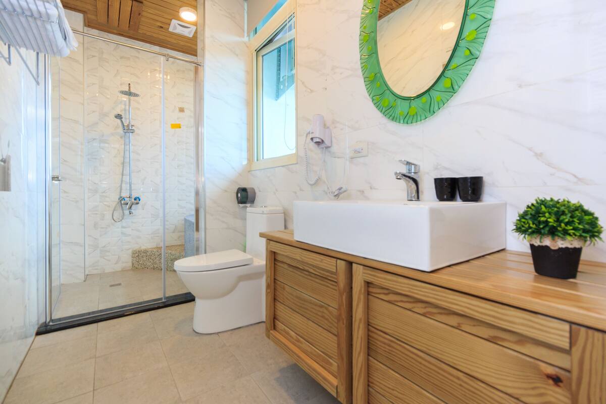 Uma casa de banho moderna com chuveiro de vidro, sanita branca, toucador de madeira com lavatório e espelho com design de folha verde.