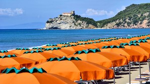 Uma praia com fileiras de guarda-sóis e cadeiras de praia laranja, um castelo no topo de uma colina e o mar ao fundo.