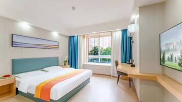Quarto de hotel com cama, escrivaninha, cadeira, janela com cortinas, um quadro na parede e iluminação no teto.